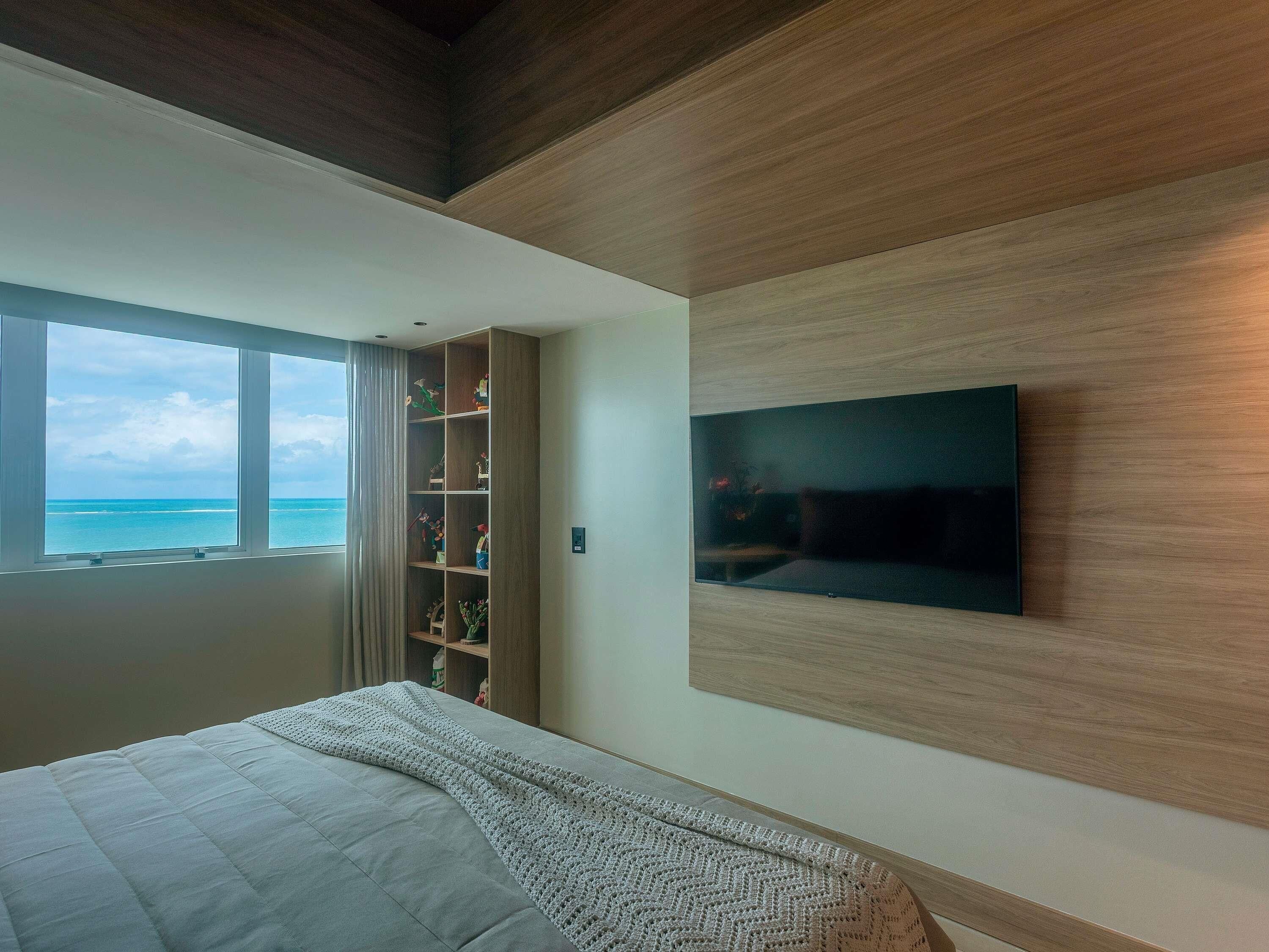 Quarto com uma janela grande, uma cama, uma TV fixada na parede e uma estante embutida.