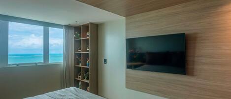 Quarto com uma janela grande, uma cama, uma TV fixada na parede e uma estante embutida.