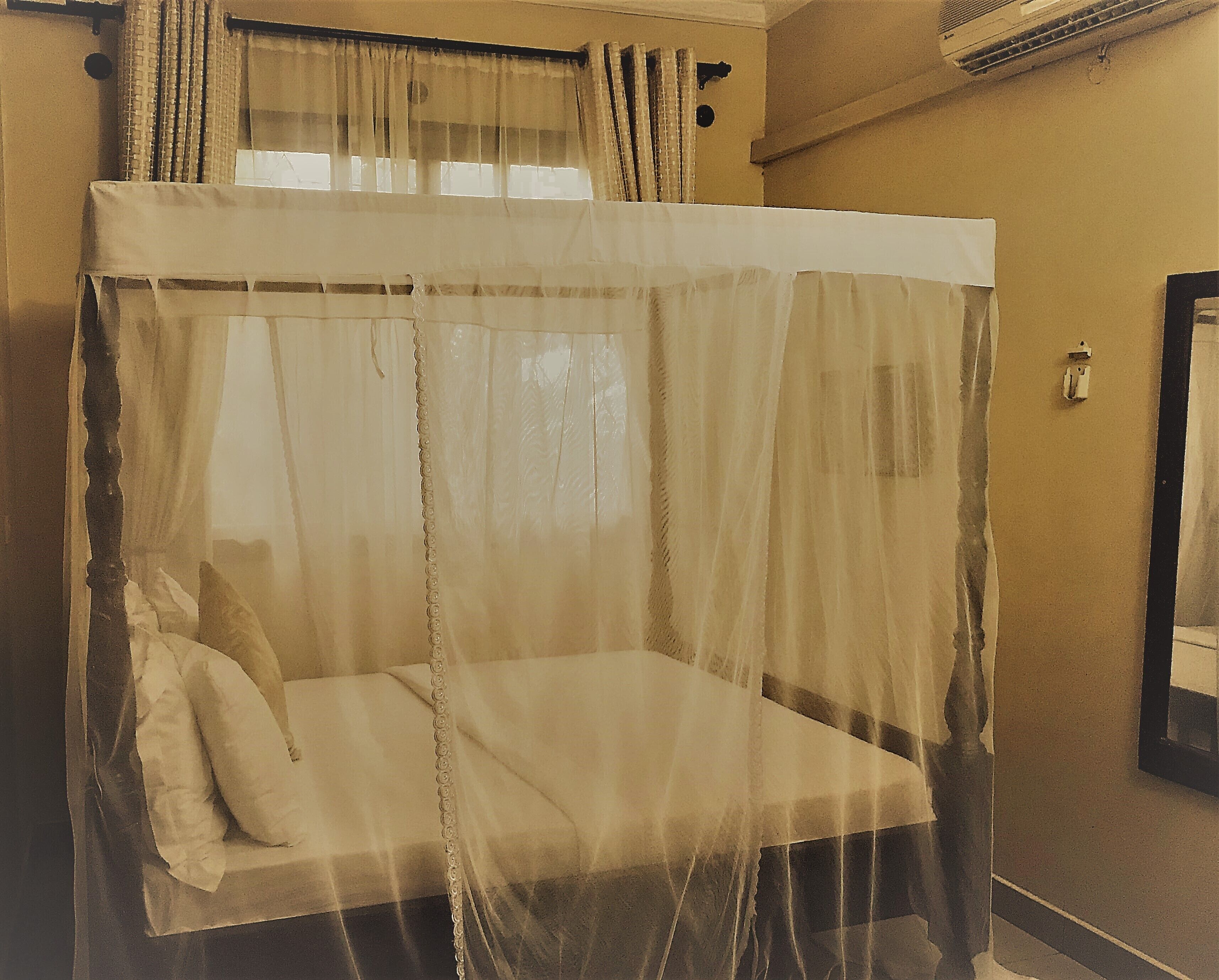 Uma cama com dossel e cortinas finas, com colcha e almofadas brancas, e cabeceira de madeira.