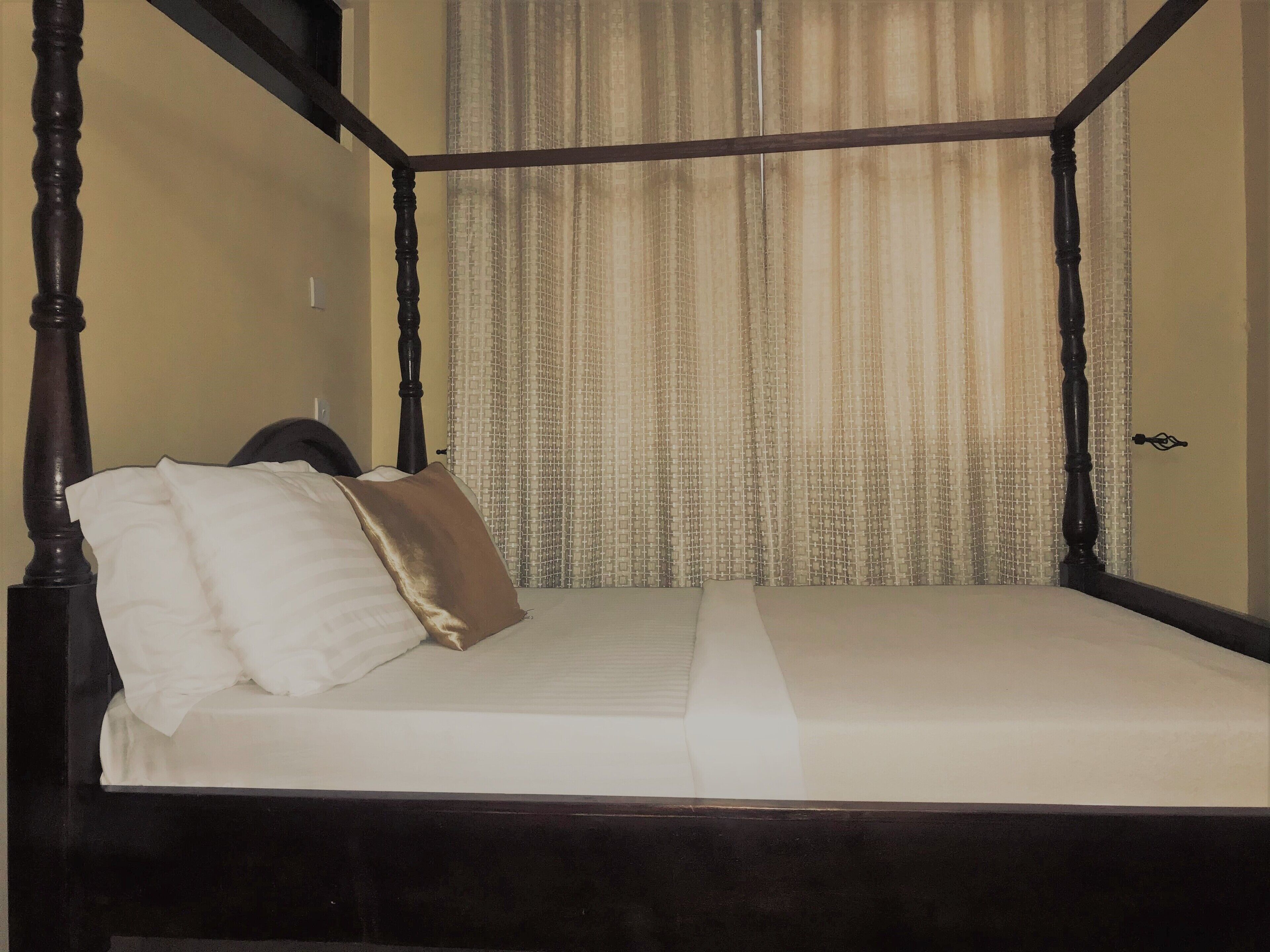 Uma cama de dossel com colchão branco e dois travesseiros, cabeceira de madeira e janela com cortina estampada.