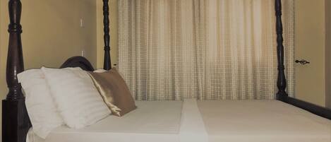 Uma cama de dossel com colchão branco e dois travesseiros, cabeceira de madeira e janela com cortina estampada.
