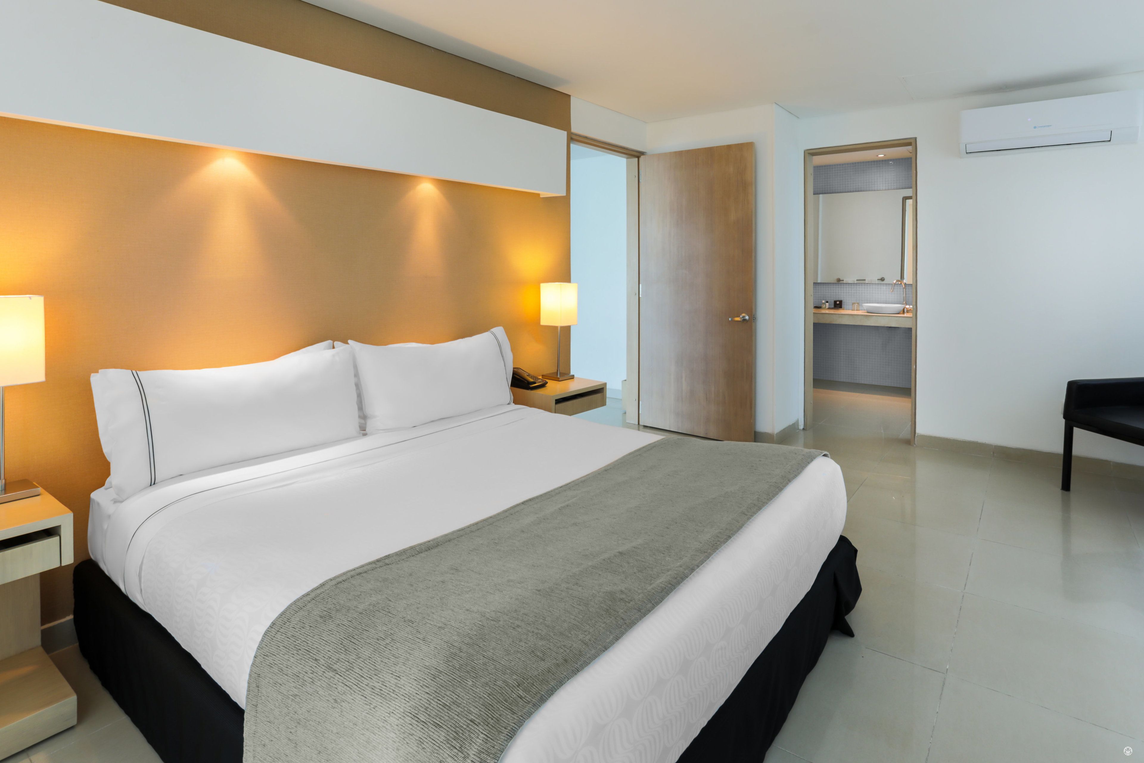 Quarto de hotel moderno com cama grande, abajures, um banco e banheiro visível por uma porta aberta.