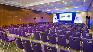 Uma sala de conferências com fileiras de cadeiras roxas, um palco com o logotipo Radisson BLU e uma parede de madeira.