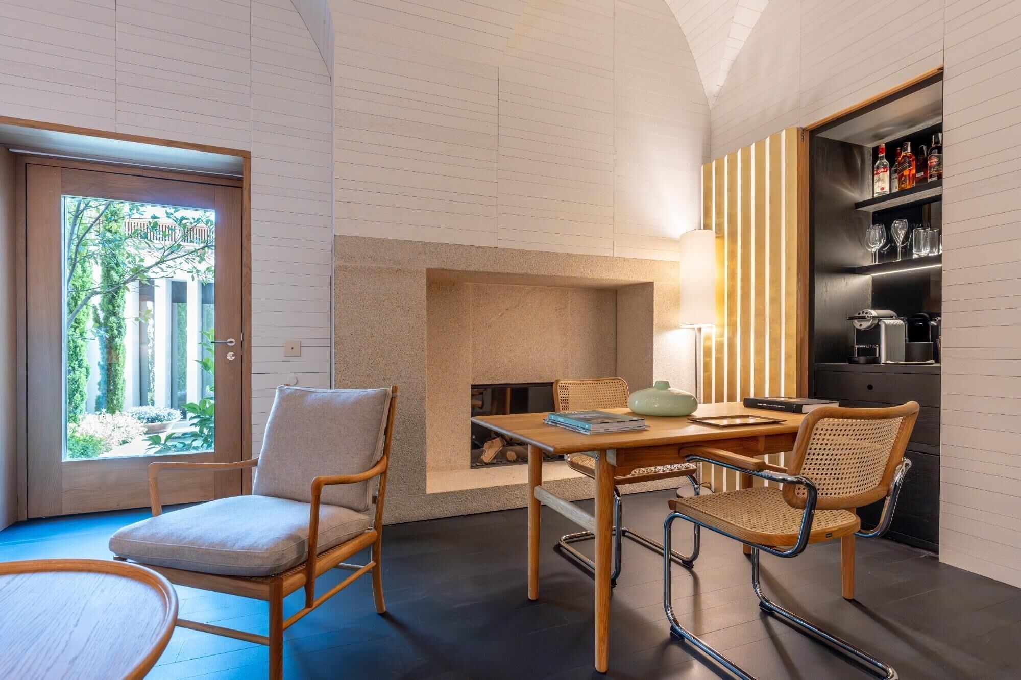 Um interior moderno com área de refeições, cadeiras, mesa, lareira e cozinha com prateleiras.