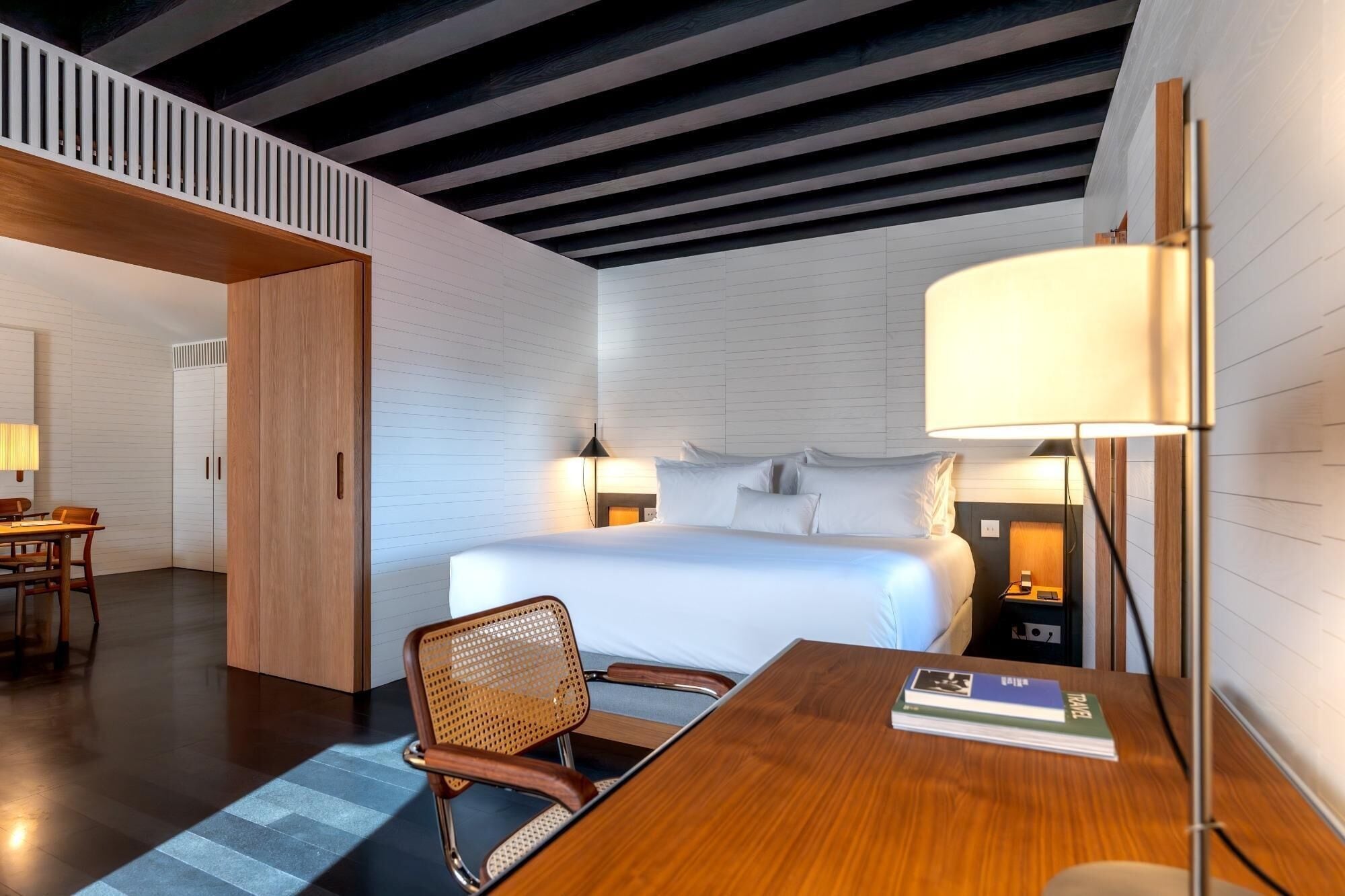 Quarto de hotel com cama, mesa de cabeceira, abajur, cadeira e escrivaninha com um livro.