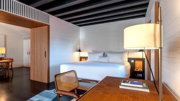 Quarto de hotel com cama, mesa de cabeceira, abajur, cadeira e escrivaninha com um livro.