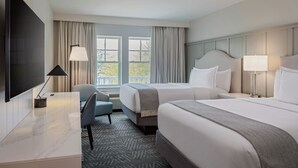 Quarto de hotel com duas camas, uma escrivaninha, uma cadeira e uma janela grande com cortinas.