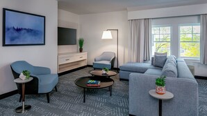 Quarto de hotel moderno com sofá, poltronas, televisão de tela plana, luminária e janela com cortinas.