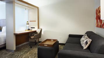 Quarto de hotel com uma cama, uma escrivaninha com cadeira, um sofá cinzento e uma mesa de apoio de madeira.