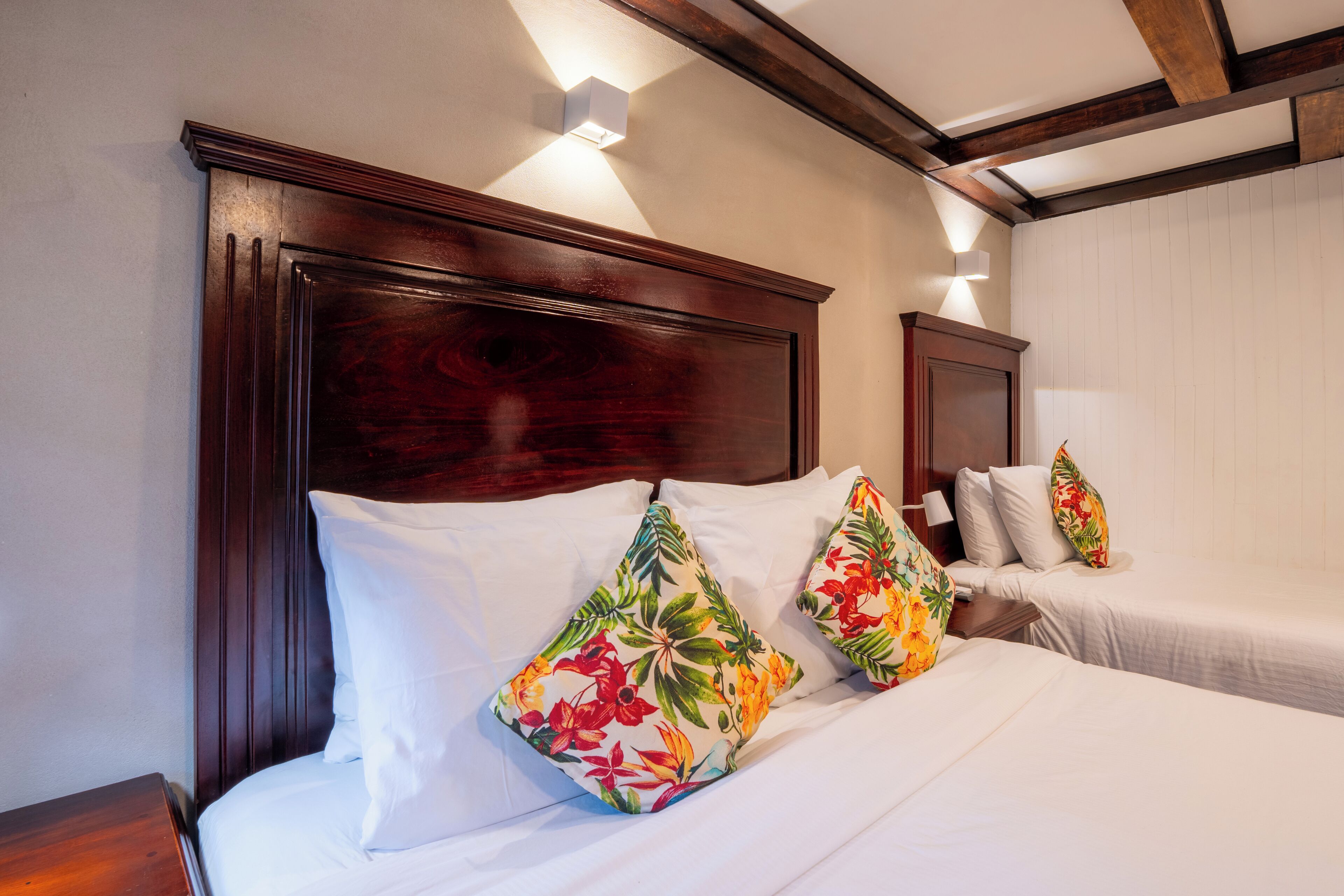 Quarto de hotel com uma grande cabeceira de madeira, duas camas com roupa de cama branca e almofadas de padrão floral.