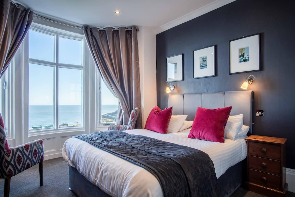 Quarto com uma cama grande, mesa de cabeceira, cadeira e vista para o mar pela janela.