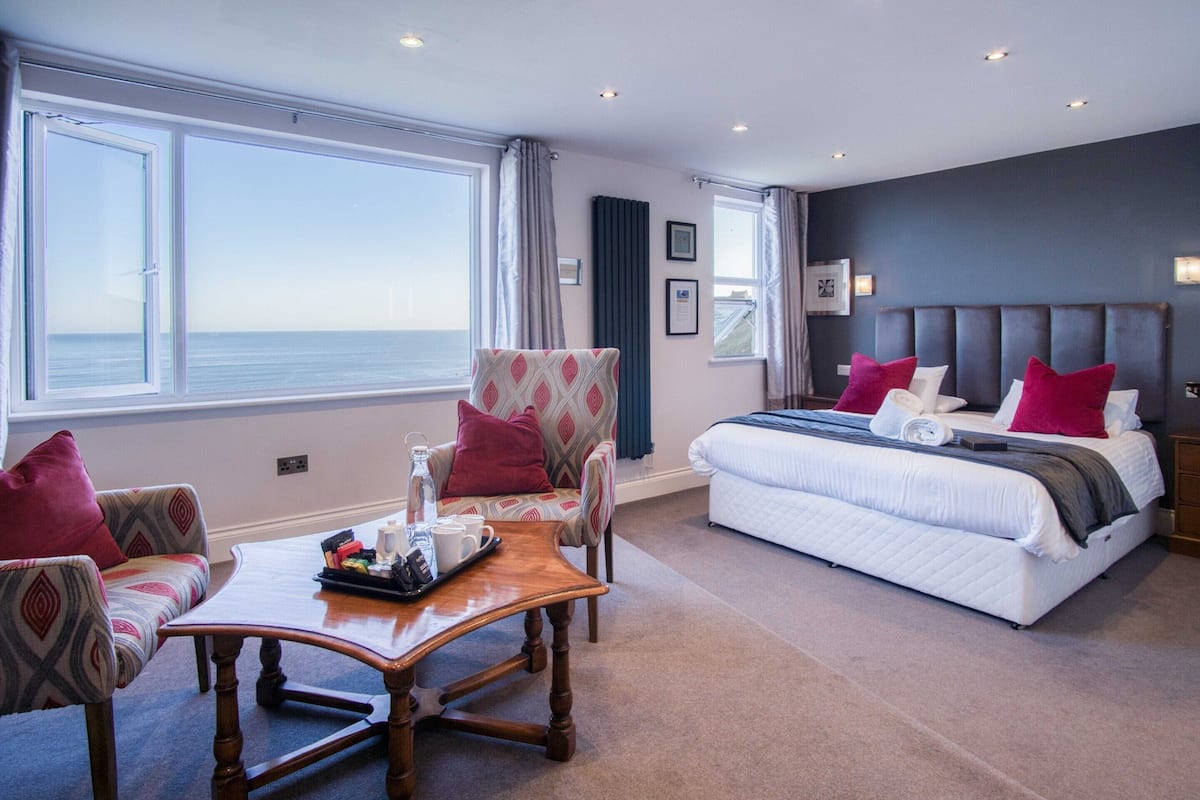 Quarto de hotel com uma janela grande, cama com almofadas, uma cadeira, mesa com objetos e piso coberto de carpete.