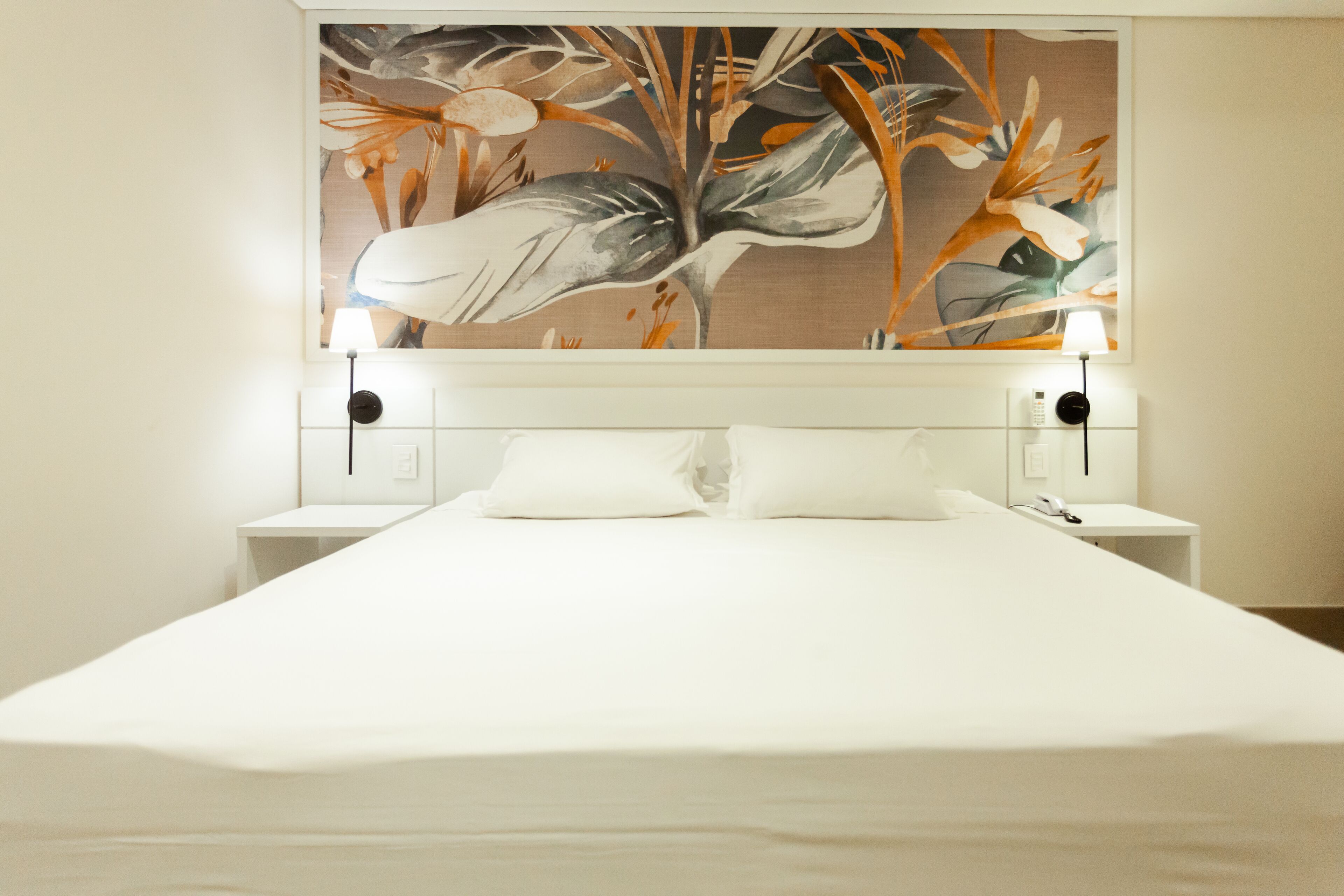 Quarto de hotel com uma cama grande, duas mesas de cabeceira e uma luminária na parede.