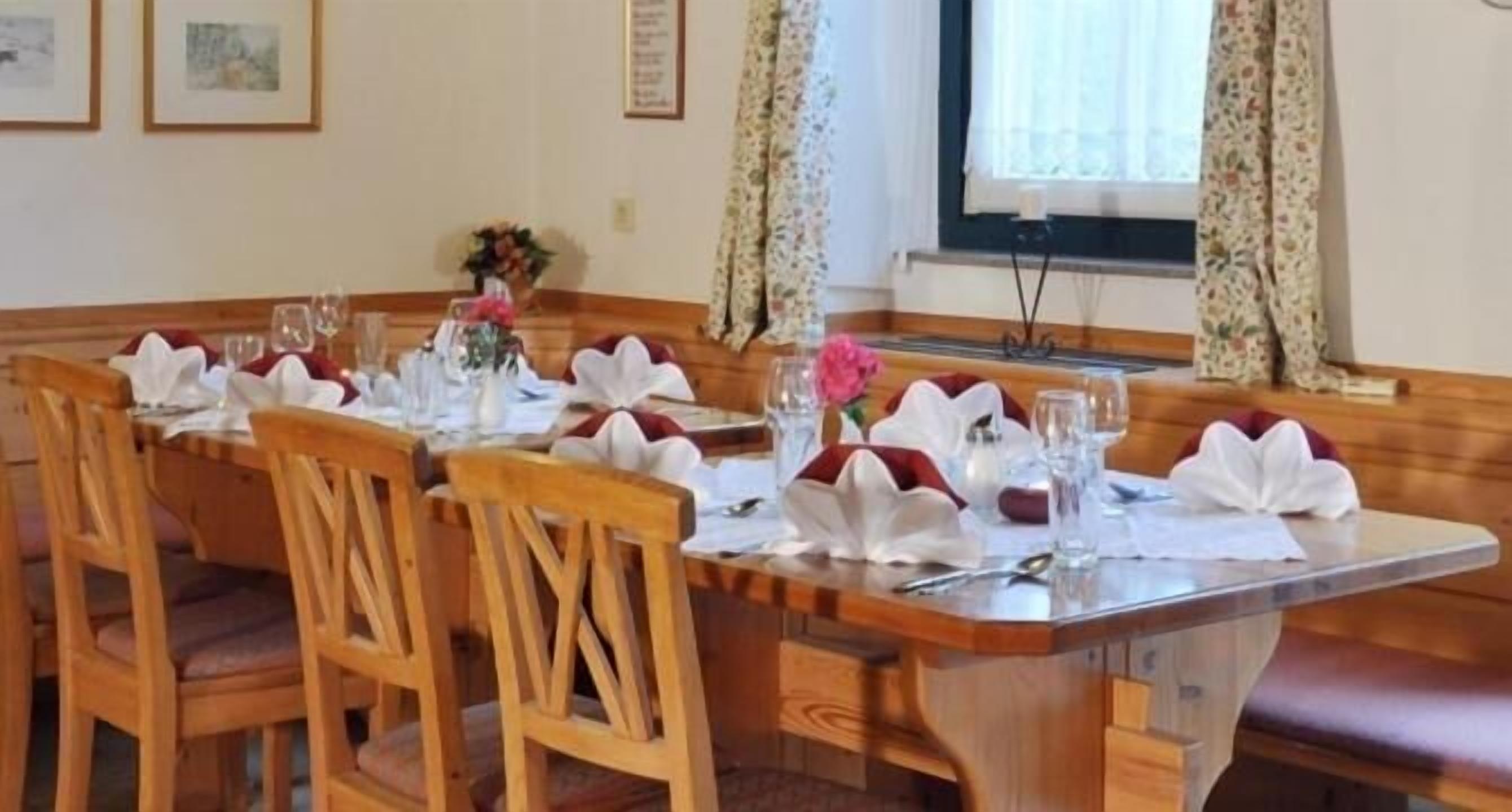 Mesa de jantar posta com toalhas brancas, guardanapos vermelhos e copos, rodeada por cadeiras de madeira.