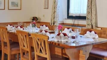 Mesa de jantar posta com toalhas brancas, guardanapos vermelhos e copos, rodeada por cadeiras de madeira.
