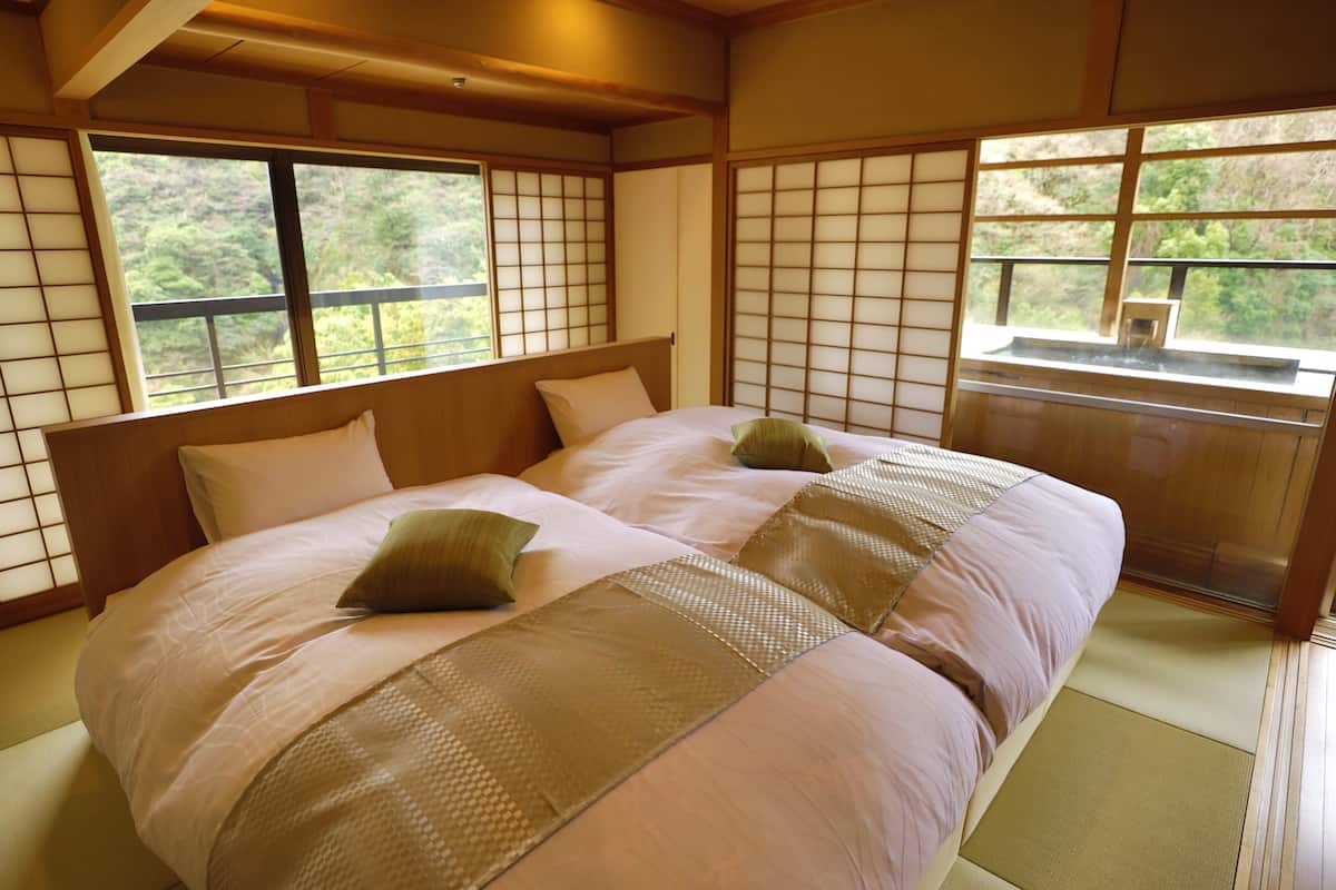Quarto japonês tradicional com tatames, uma cama grande com almofadas e vista para a floresta através de grandes janelas.