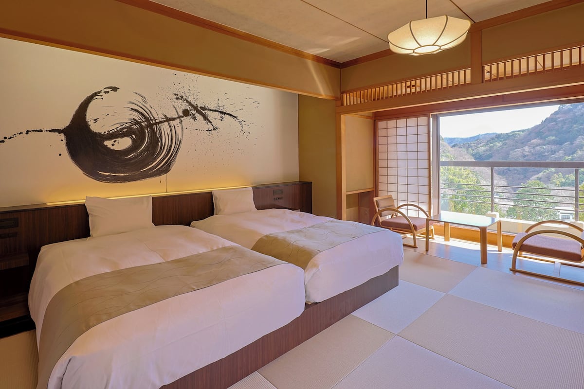 Quarto de hotel com duas camas, uma pintura grande na parede, varanda com cadeiras e vista para as montanhas.