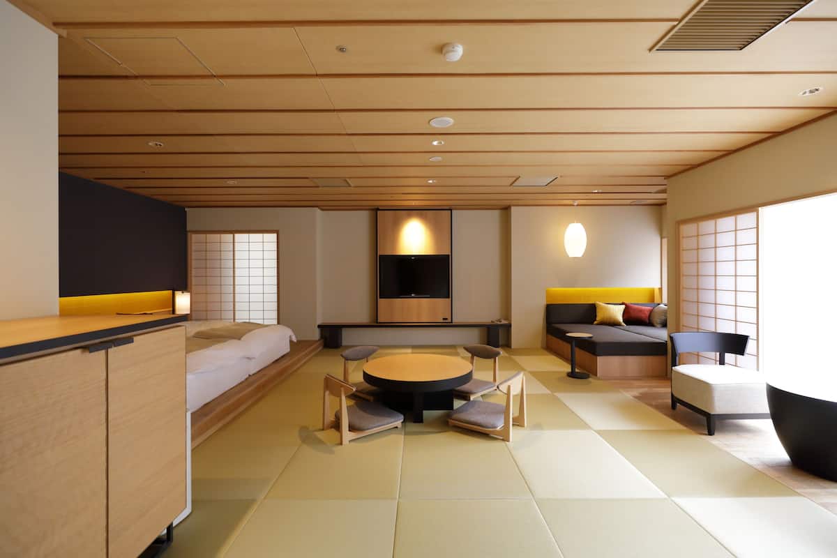 Quarto em estilo japonês tradicional, com tatames, uma mesa baixa com cadeiras, um sofá e uma banheira grande preta.