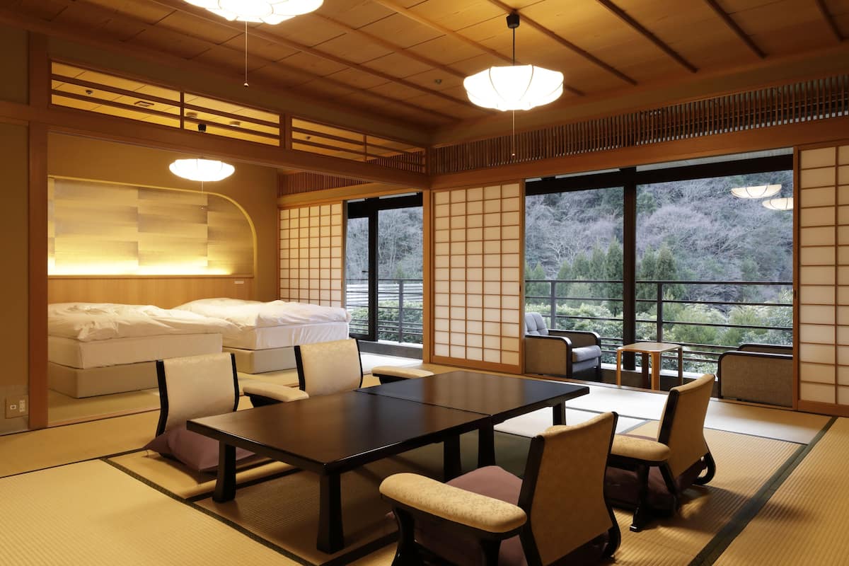 Um quarto tradicional japonês com tatames, uma mesa baixa com cadeiras e uma cama com roupa de cama branca.