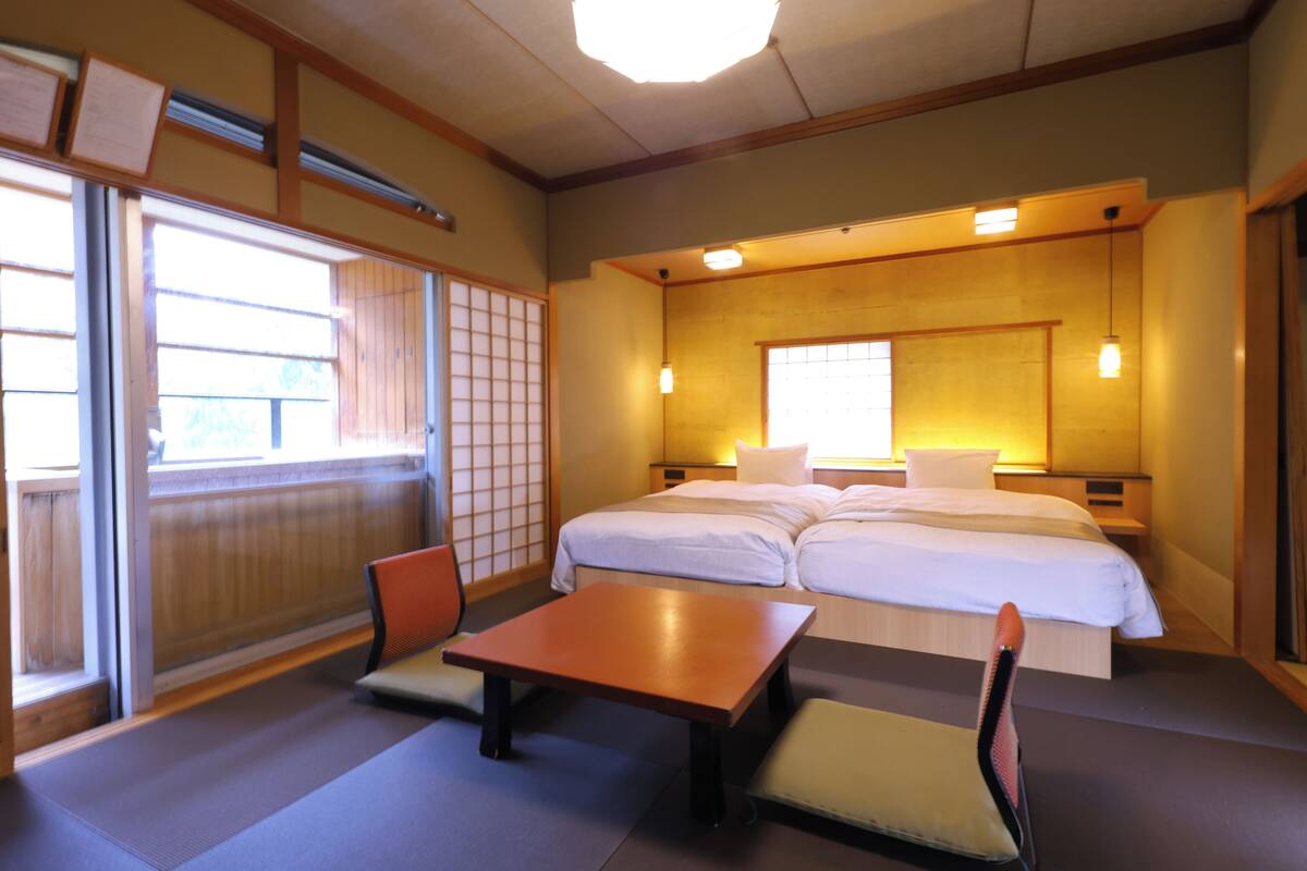 Um quarto japonês tradicional, com piso de tatame, uma mesa baixa, duas cadeiras e uma cama com duas almofadas.