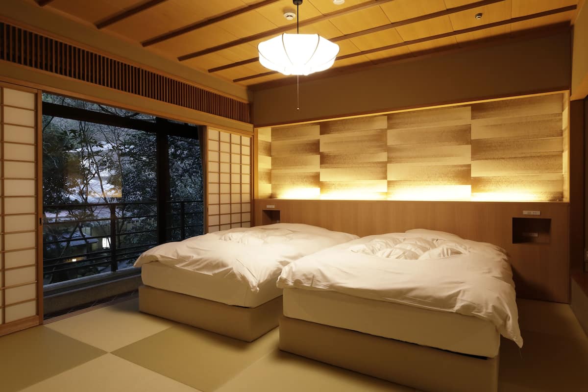 Quarto tradicional japonês com tatames, um painel shoji e uma grande janela com vista para árvores.