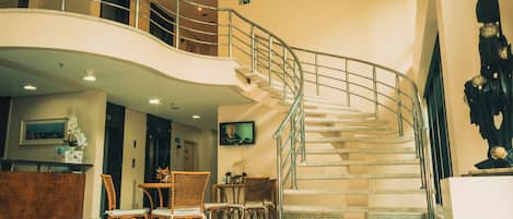 Um saguão de hotel com uma escadaria curva, um balcão de recepção, cadeiras de vime e uma tela de televisão.