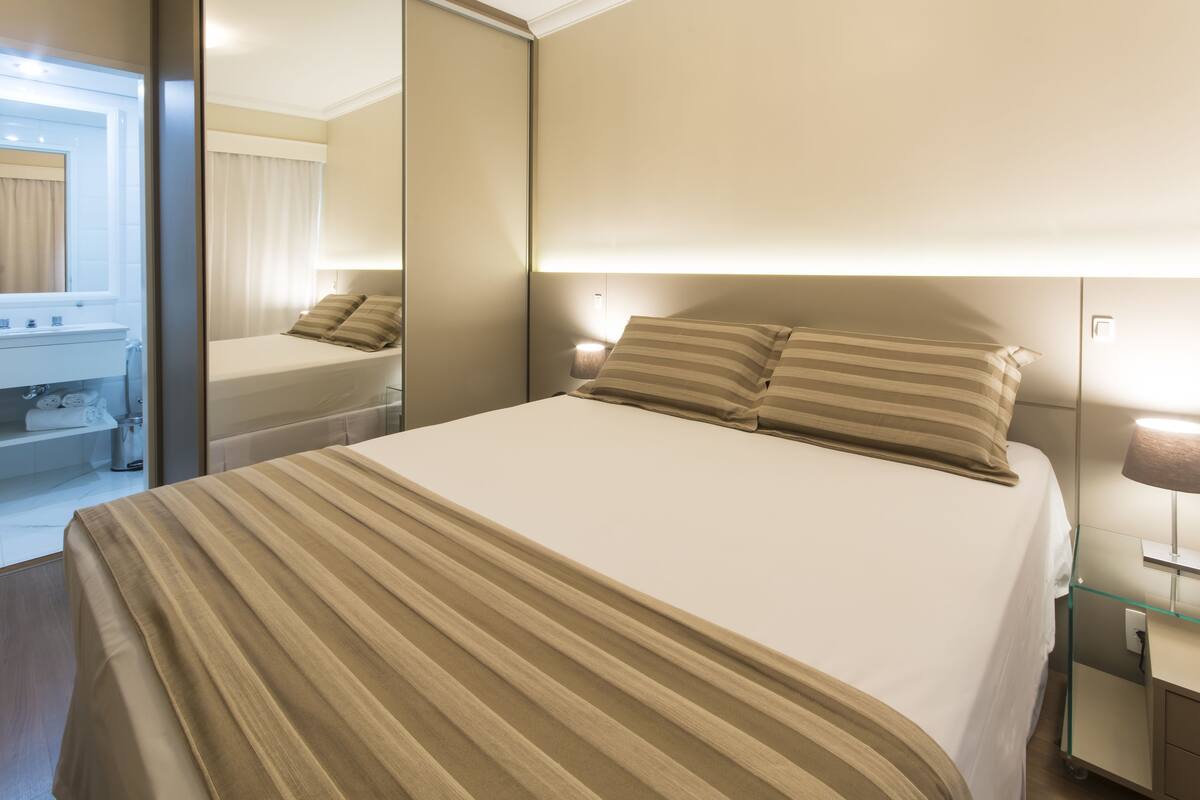 Quarto de hotel com uma cama grande, mesa de cabeceira, espelho e banheiro com pia e porta-toalhas.