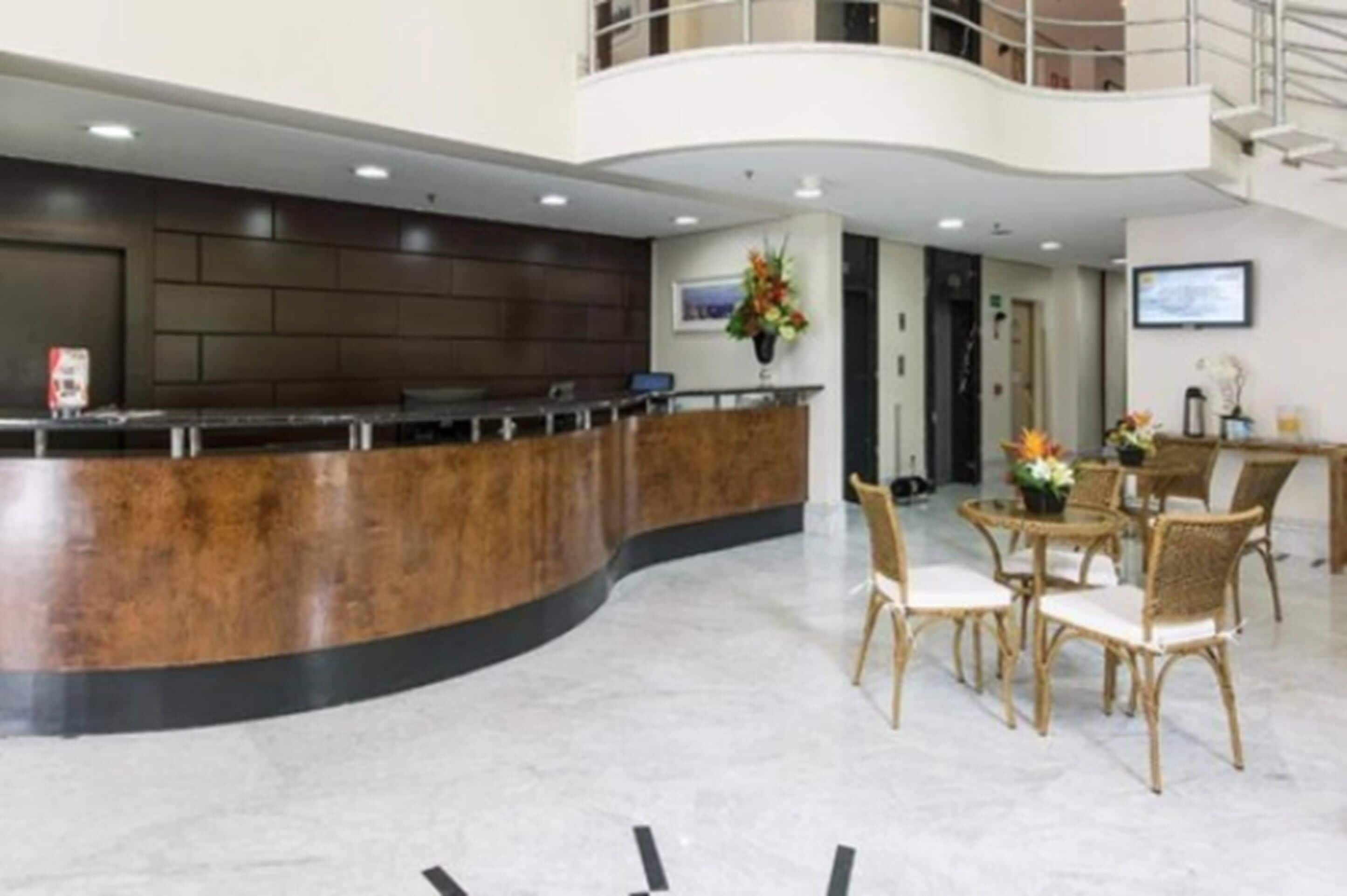 Uma área de recepção de hotel com um balcão curvo, um espaço de estar com cadeiras de vime e uma escadaria que leva a um andar superior.