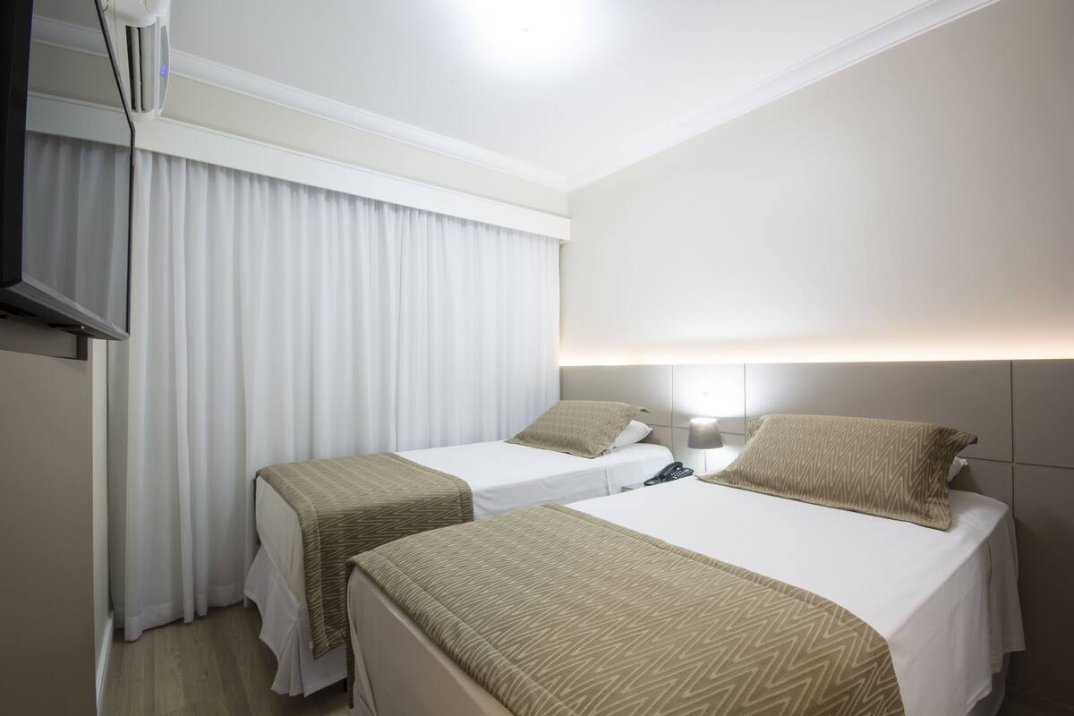 Quarto de hotel com duas camas, cada uma com colcha estampada e almofadas, uma mesa de cabeceira com abajur, e uma janela grande com cortinas brancas.