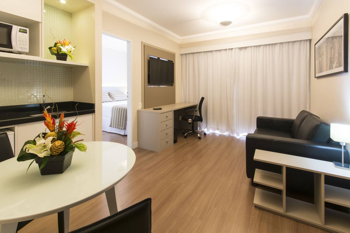Quarto de hotel moderno com área de jantar, sala de estar, escrivaninha com cadeira, televisão de tela plana e uma grande janela com cortinas.