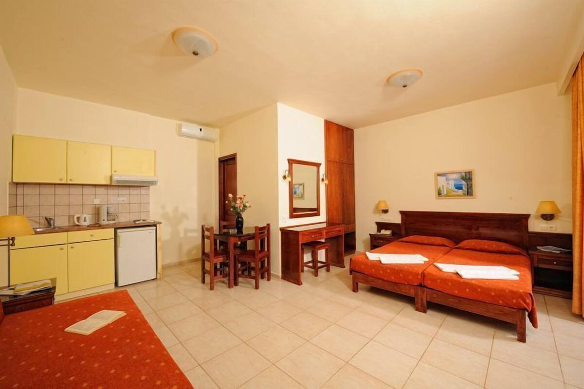 Quarto de hotel compacto com cozinha compacta, uma cama de solteiro com duas almofadas, uma escrivaninha com cadeira e uma pequena mesa de cabeceira.