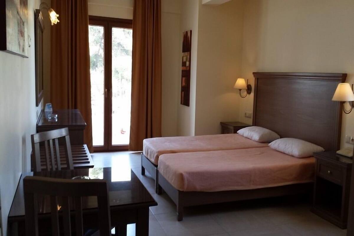 Quarto de hotel com duas camas, uma escrivaninha com cadeira, uma mesinha e uma janela com cortinas.