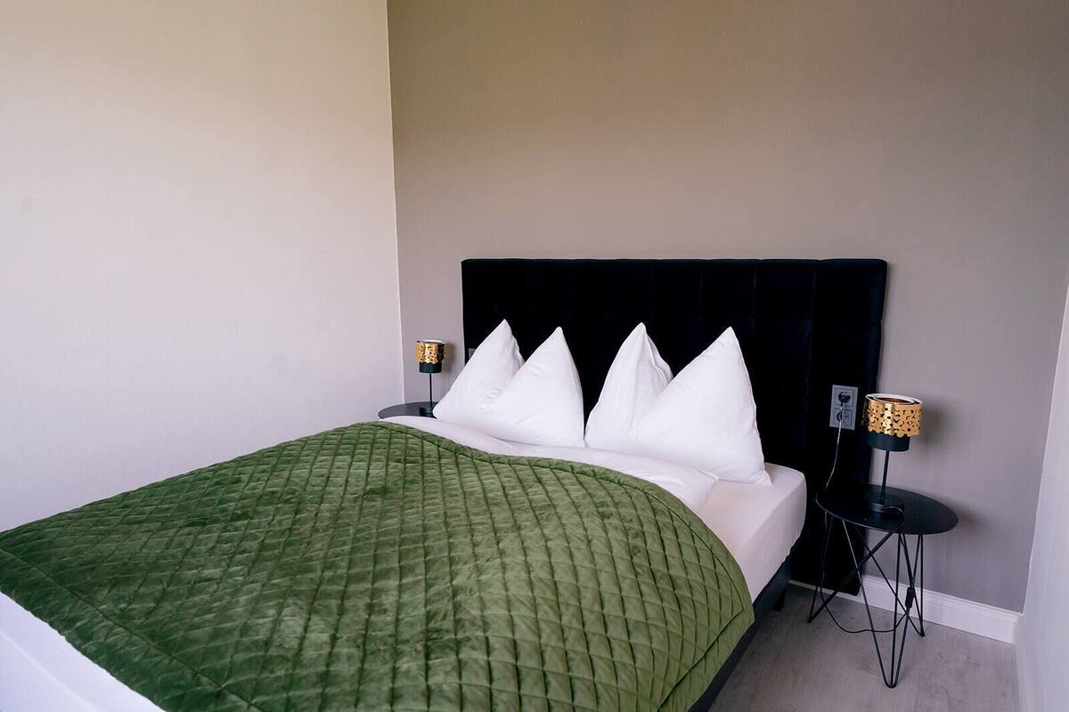 Uma cama bem arrumada com um edredom verde, almofadas brancas e uma cabeceira preta. Há duas mesas de cabeceira com abajures.