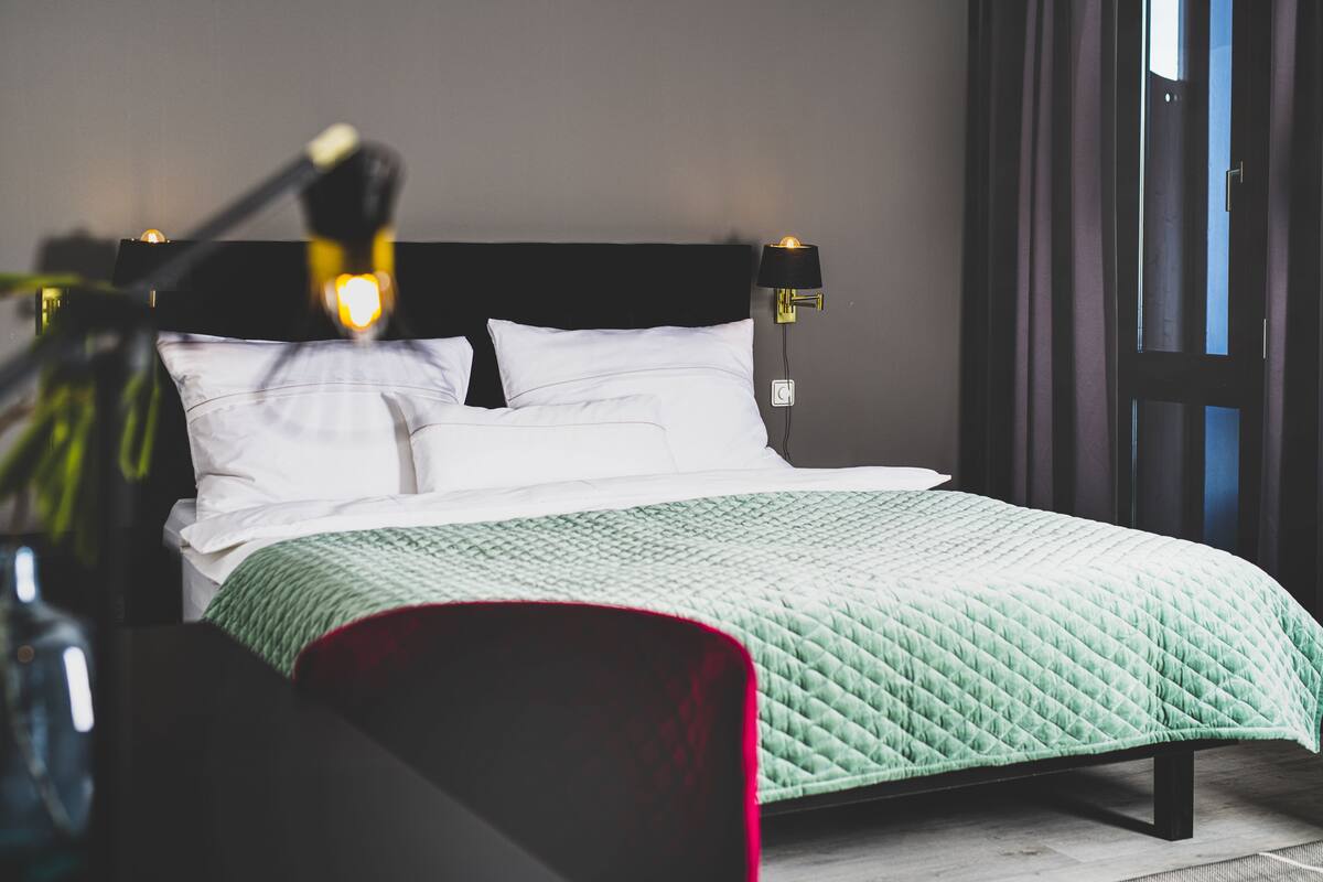 Cama bem arrumada com almofadas brancas e um edredom verde, luminária de cabeceira e vista para uma janela com cortinas.
