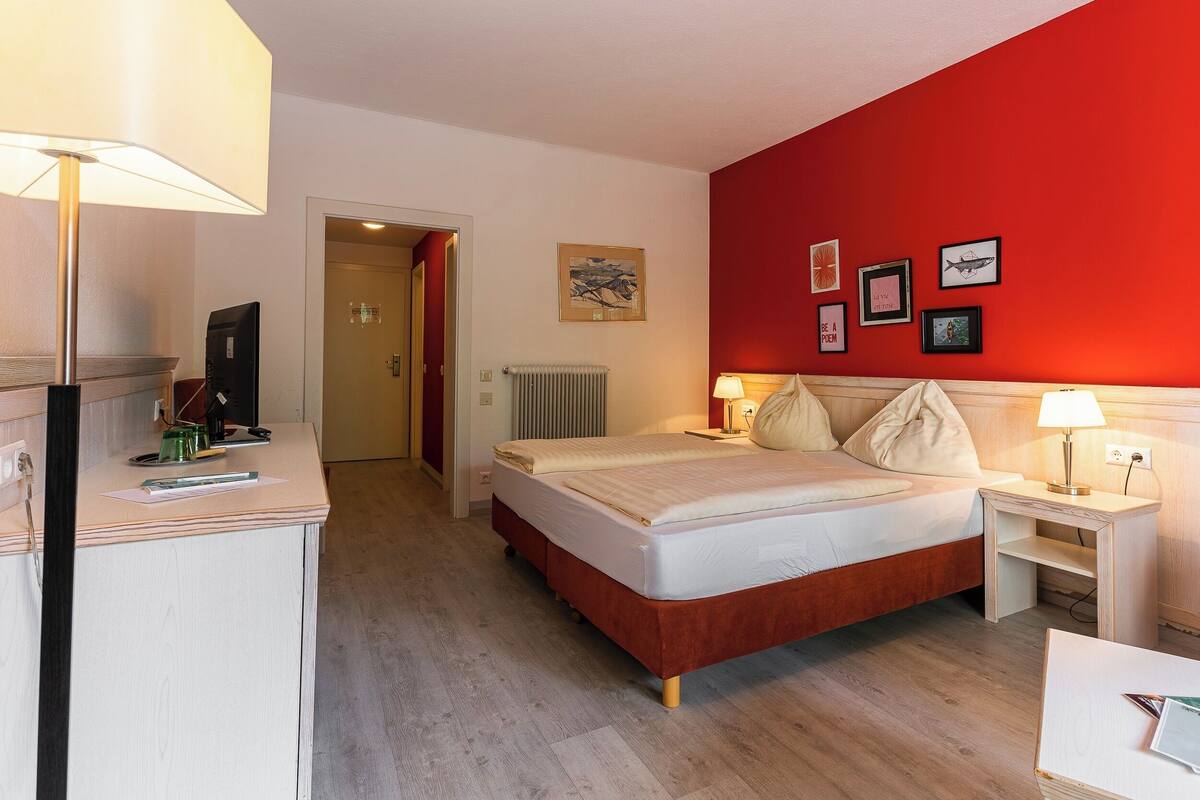 Quarto de hotel com cama, mesas de cabeceira, escrivaninha e luminária.