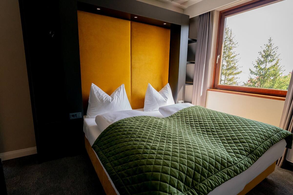Quarto de hotel com cama, cabeceira amarela, almofadas brancas e colcha verde acolchoada.