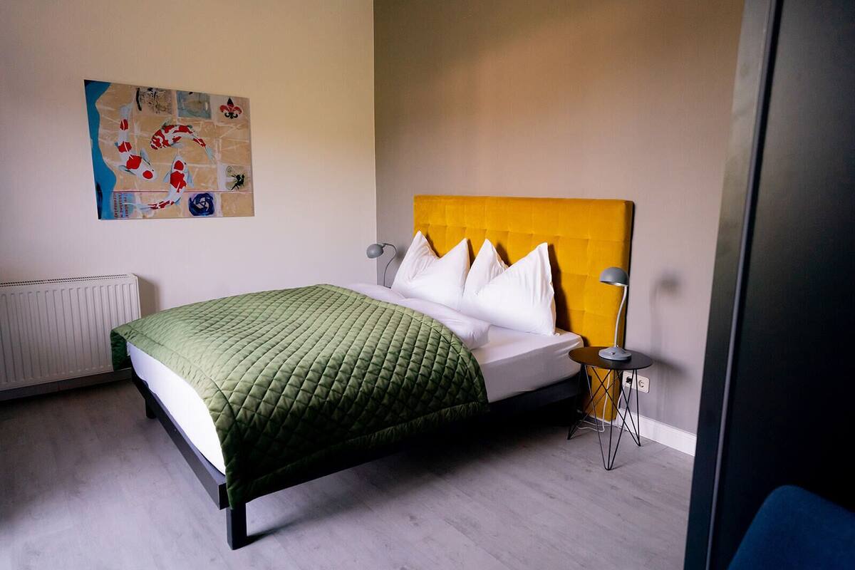Cama bem arrumada com colcha verde, almofadas brancas e cabeceira amarela em quarto moderno, com radiador e uma obra de arte na parede.