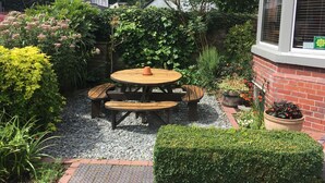 Um pátio com jardim, mesa e bancos de madeira, cercado por plantas diversas e um caminho de cascalho.