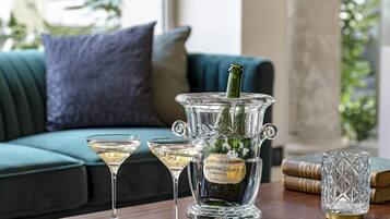 Um jarro de vidro e dois copos de martini sobre uma mesa de madeira, com um sofá verde e livros ao fundo.