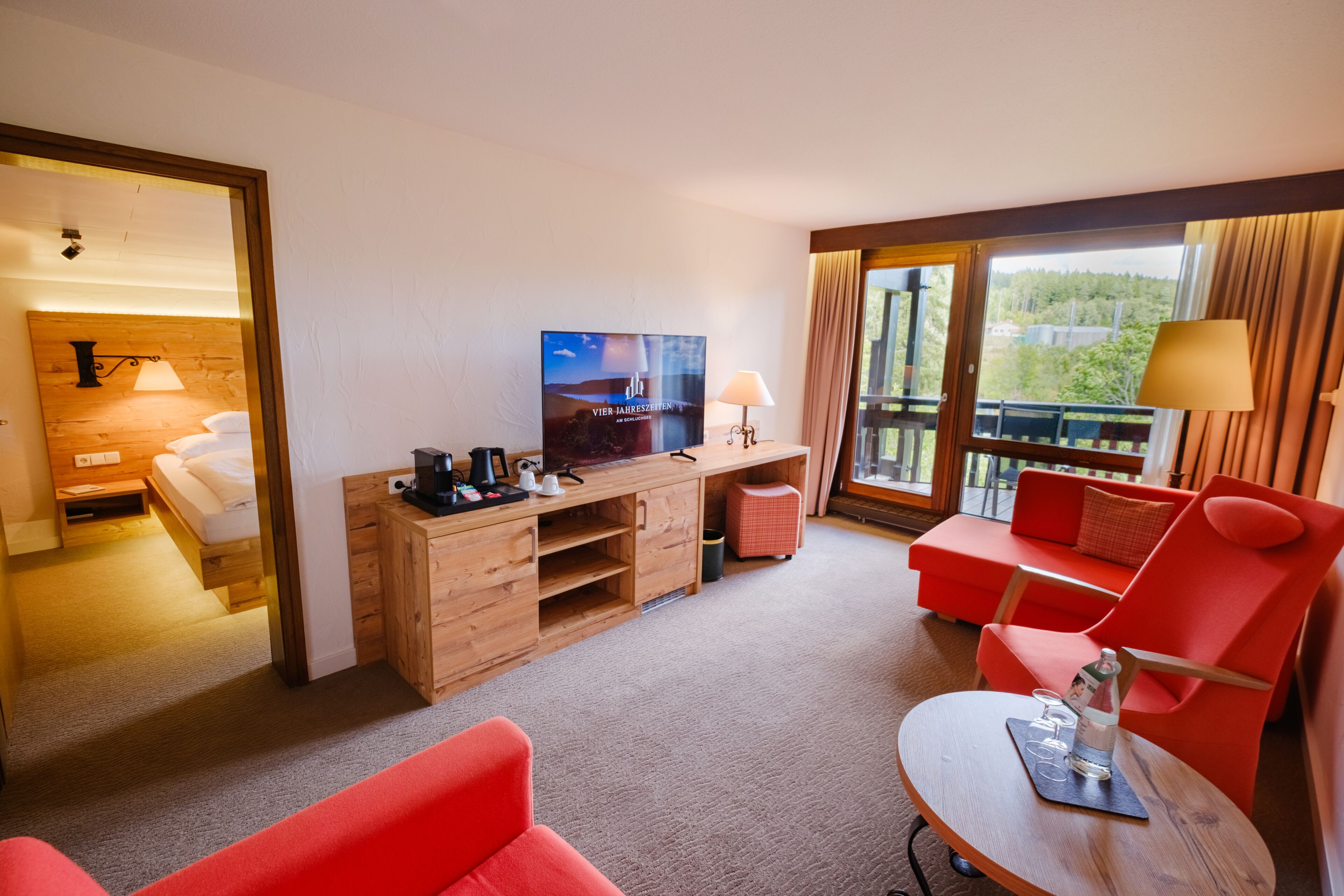 Quarto de hotel com televisão, sofá, cadeira vermelha, mesa pequena e janela com vista.