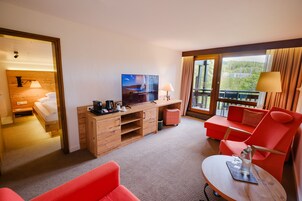 Quarto de hotel com televisão, sofá, cadeira vermelha, mesa pequena e janela com vista.