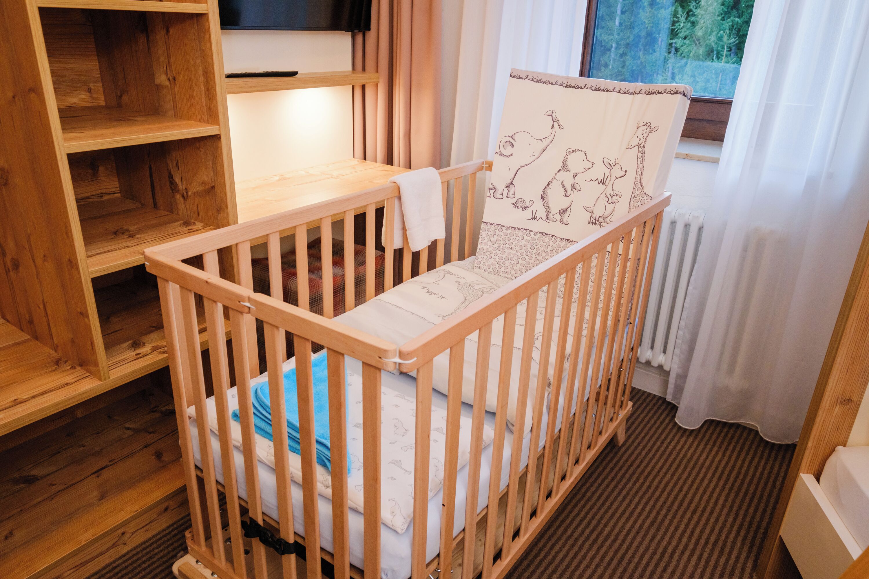Um berço de madeira com travesseiro estampado, uma estante de madeira e vista para uma janela com cortinas.