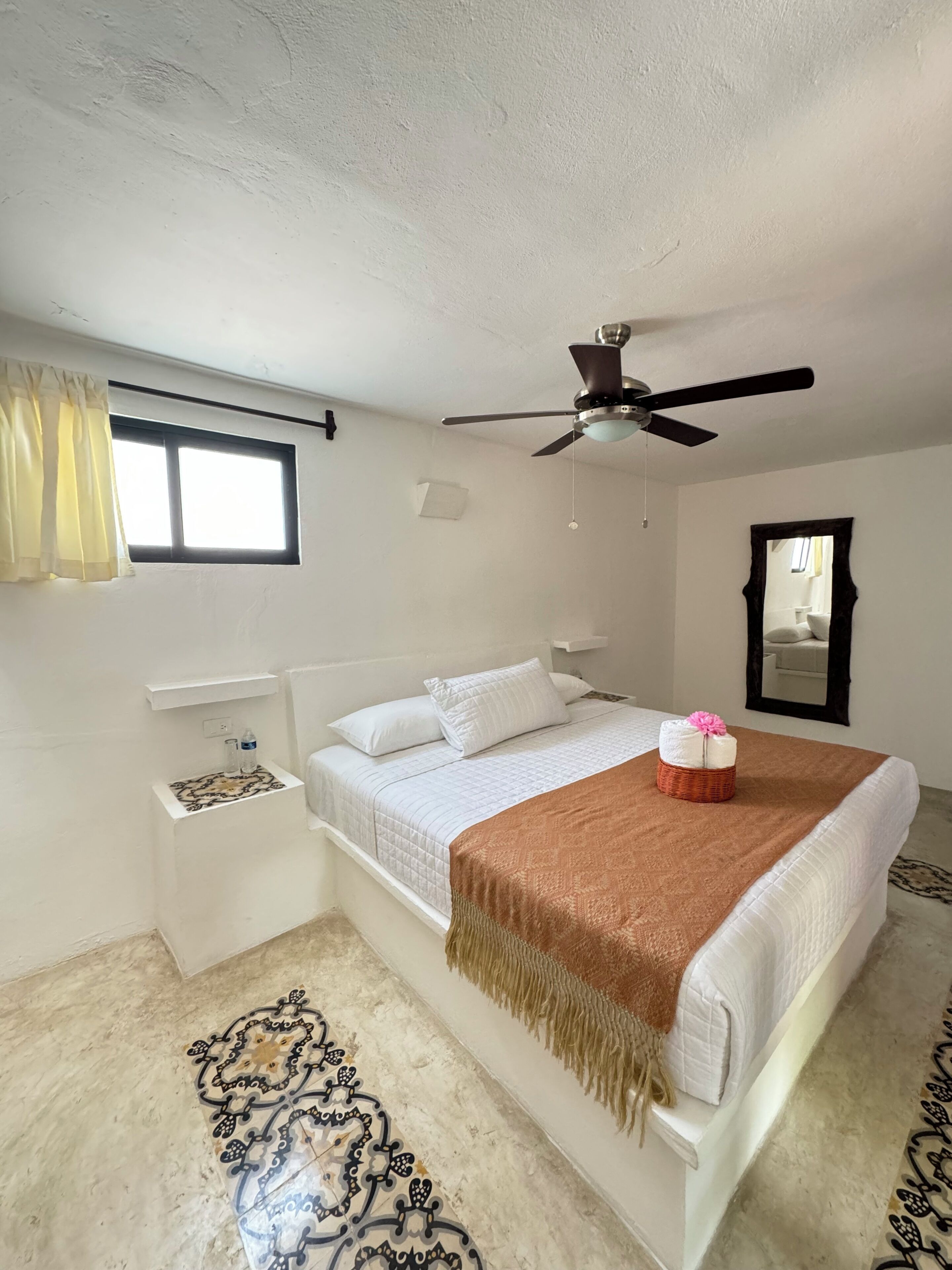 Quarto com cama, ventoinha no teto, janela com cortinas, mesa de cabeceira e tapete estampado.