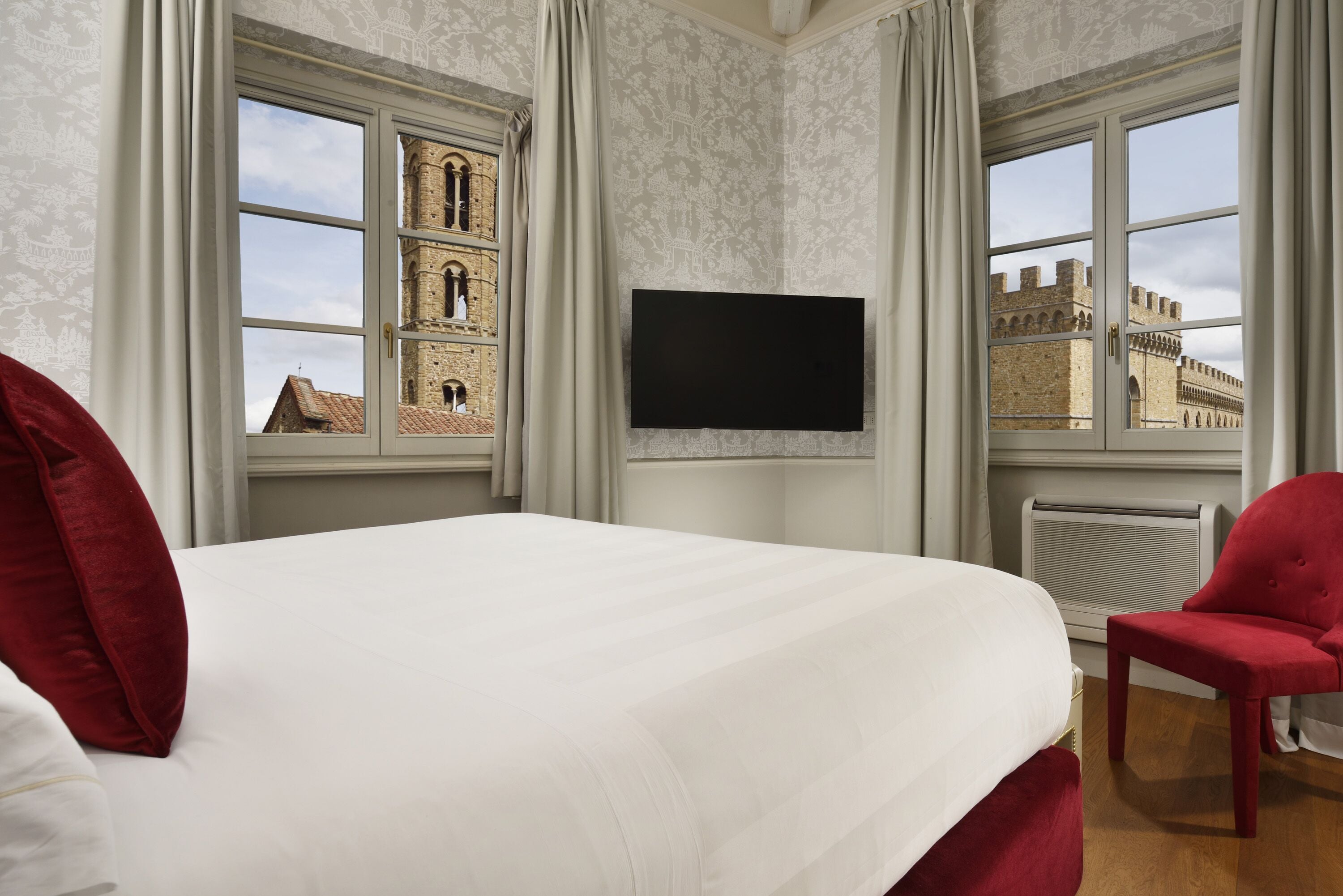 Quarto de hotel com uma cama grande, uma cadeira vermelha e uma televisão fixada na parede.