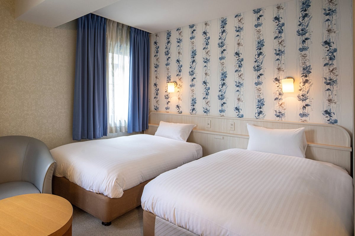 Quarto de hotel com duas camas, uma cadeira, uma mesa redonda e papel de parede floral.