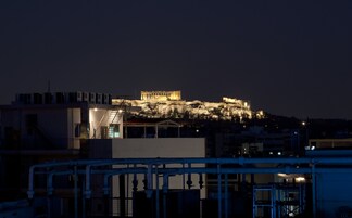 Vista noturna de uma paisagem urbana com um antigo anfiteatro iluminado em destaque numa colina, edifícios modernos em primeiro plano e um corpo de água com uma ponte.