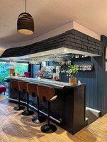 Uma área de bar com balcão preto, banquetas de madeira e um lustre suspenso.