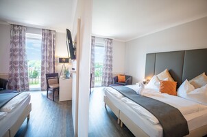 Quarto de hotel com uma cama grande, uma escrivaninha e uma cadeira. Há uma televisão na parede e uma janela com cortinas.