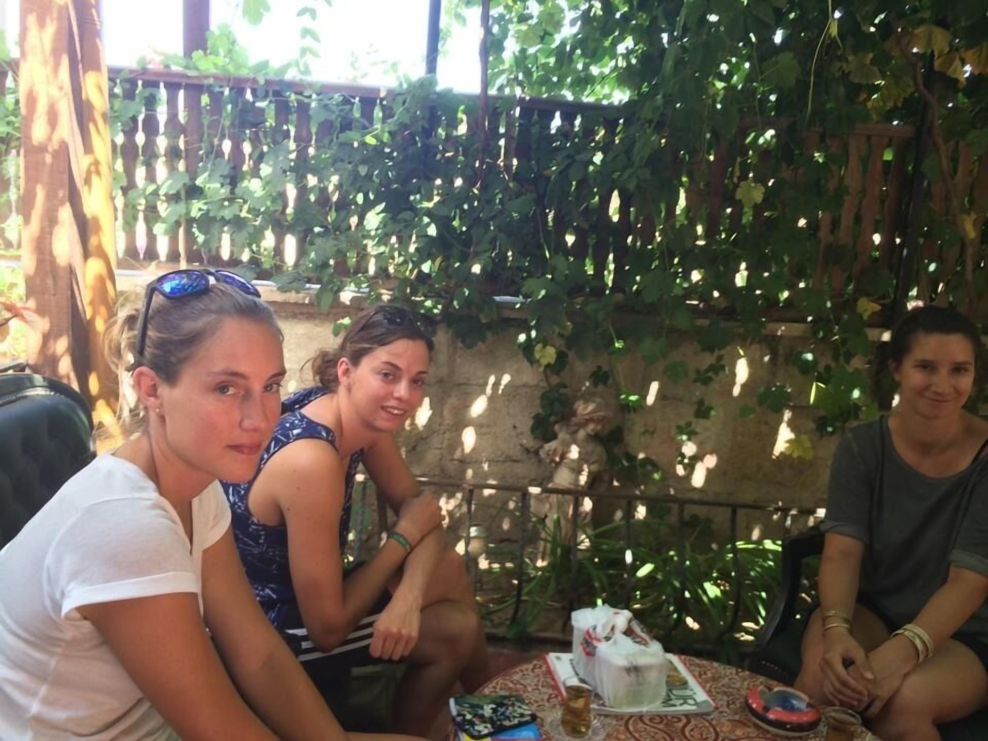 Um grupo de pessoas sentadas numa mesa ao ar livre, com bebida e bolo, rodeadas por vegetação e uma cerca de madeira.