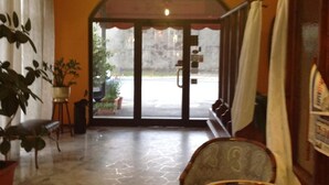 Um hall de hotel com ventoinha no teto, chão de pedra, uma cadeira, uma mesa pequena e vista através das portas de vidro.